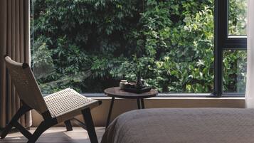 Quarto com uma cama, uma cadeira de vime, uma pequena mesa com uma planta e vista para um verde exuberante pela janela.