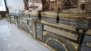 Exposição de relógios ornamentados em molduras douradas, em um quarto moderno e bem iluminado, com design geométrico no teto.