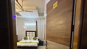 Quarto de hotel com cama, porta de madeira com um número, ventilador de teto e banheiro com vaso sanitário.