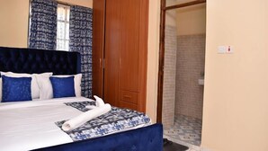 Quarto de hotel com uma cama, um guarda-roupa de madeira e um banheiro visível através de uma porta de vidro.