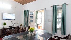 Uma área de refeições com mesa de vidro, cadeiras, TV fixada na parede e porta que dá para outro cômodo.