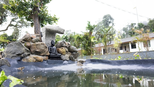 Um lago tranquilo com uma estátua de Buda, cercado por pedras e vegetação, uma casa ao fundo, e céu limpo.