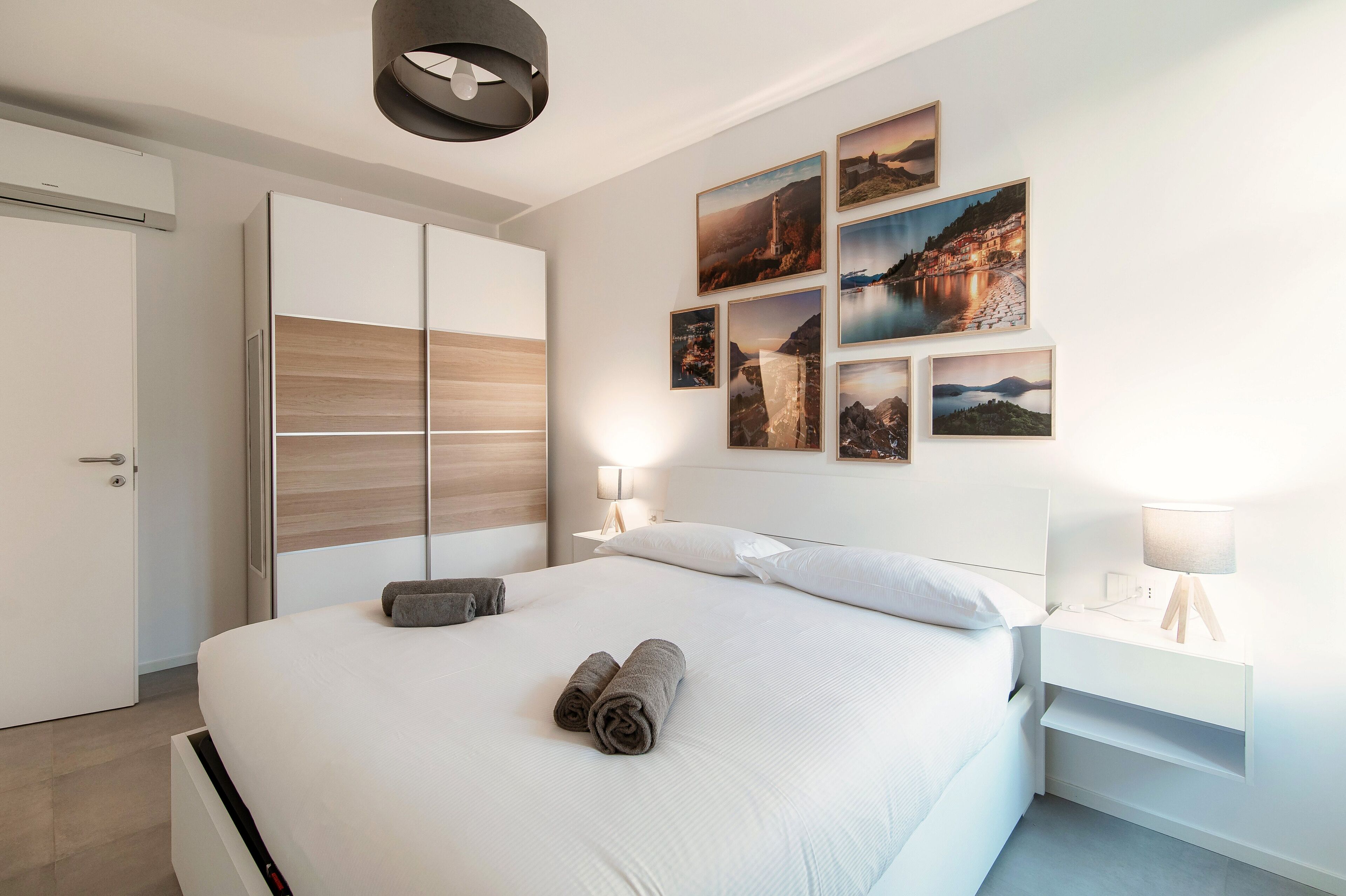 Quarto de hotel moderno com uma cama grande, um guarda-roupa, uma mesa de cabeceira com uma luminária e uma parede decorada com fotografias de paisagens.
