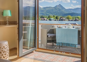 Uma varanda com mesa posta para chá, uma cadeira de vime e vista para um lago com montanhas ao fundo.