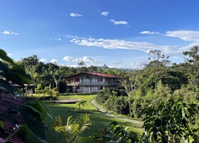 Um edifício de dois andares com telhado vermelho, rodeado por vegetação exuberante e um céu azul límpido.