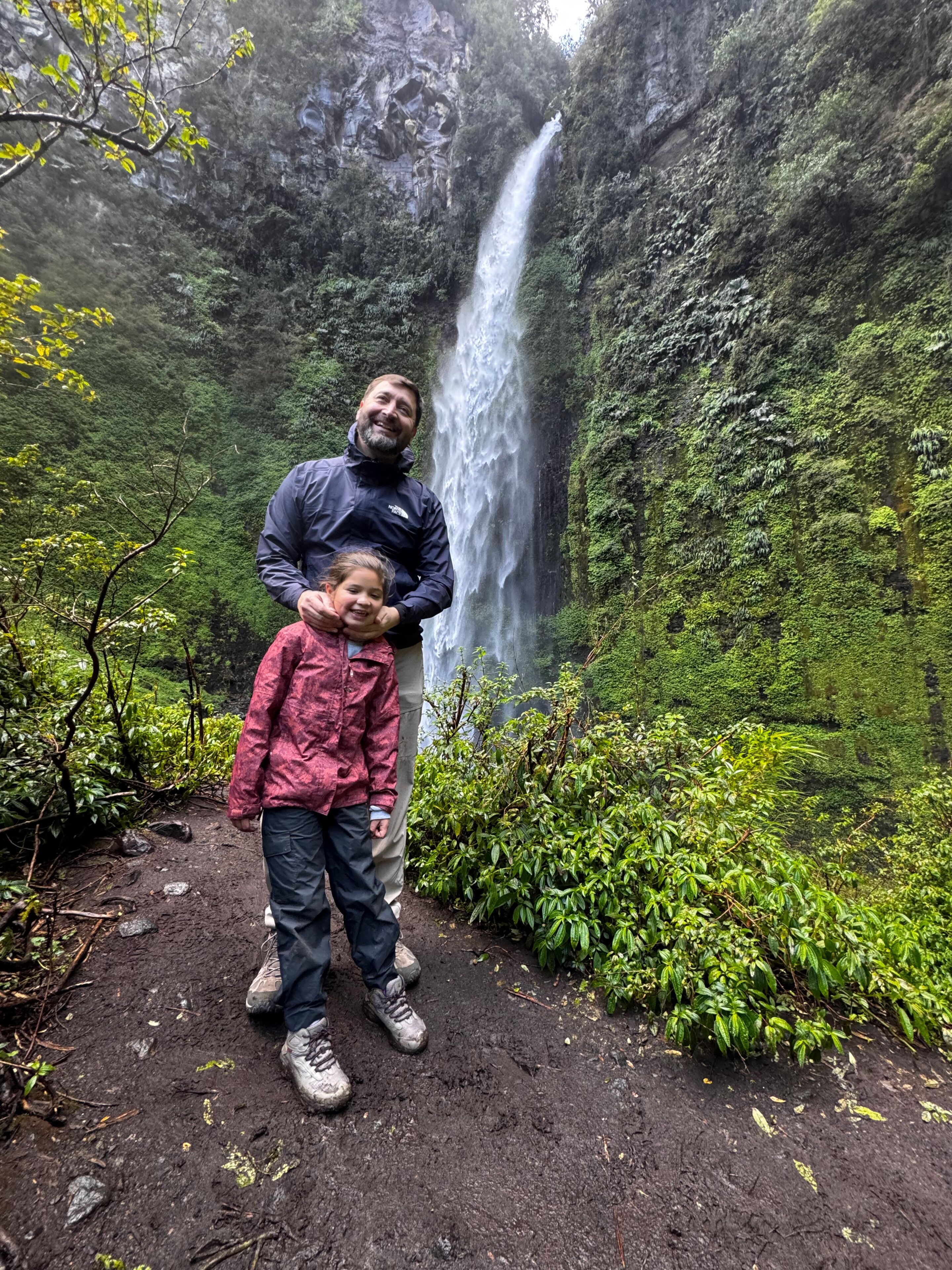 Um homem e uma criança em frente a uma cachoeira, com vegetação exuberante e penhascos rochosos ao fundo.