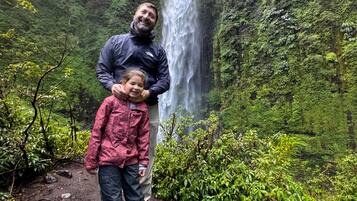 Um homem e uma criança em frente a uma cachoeira, com vegetação exuberante e penhascos rochosos ao fundo.