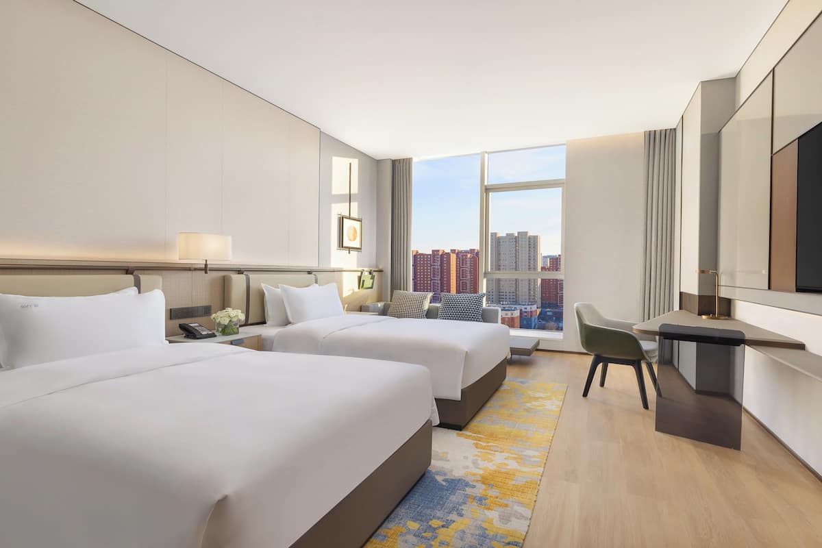 Quarto de hotel com duas camas, uma escrivaninha e uma janela grande com vista para a cidade.