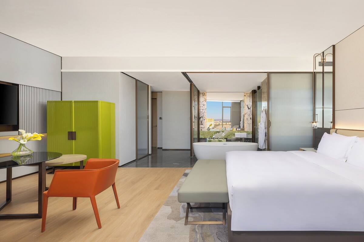 Quarto de hotel moderno com uma cama grande, uma escrivaninha com cadeira, um armário verde e vista para a paisagem urbana.