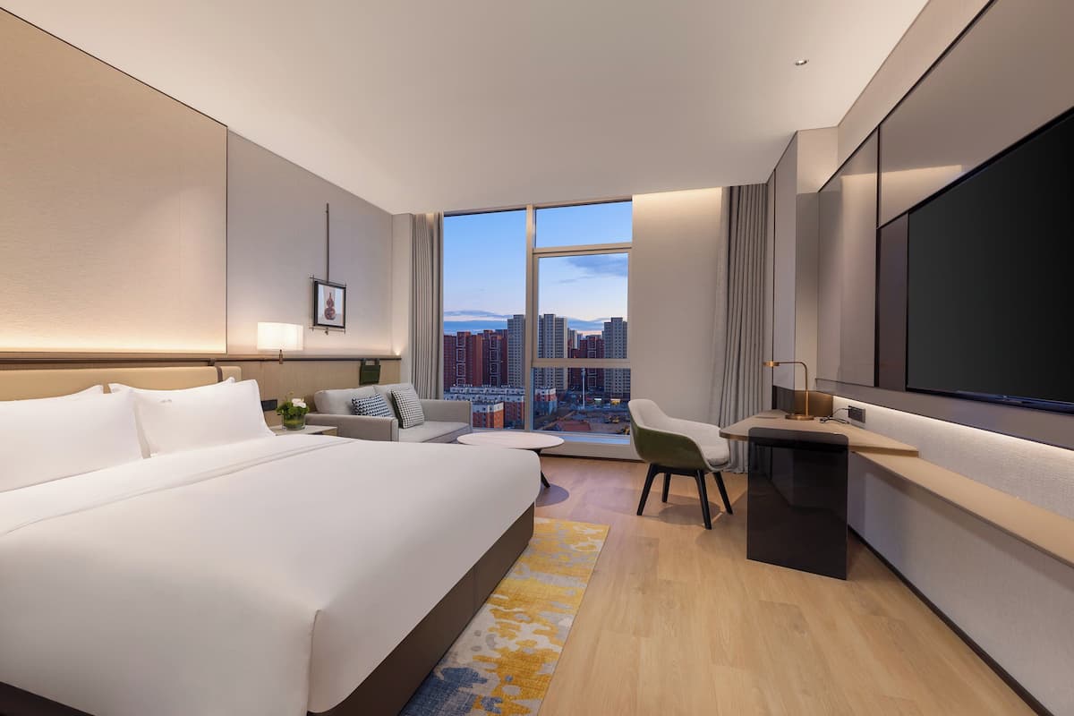 Quarto de hotel moderno com uma cama grande, uma mesa, uma cadeira e uma janela ampla com vista para a cidade.