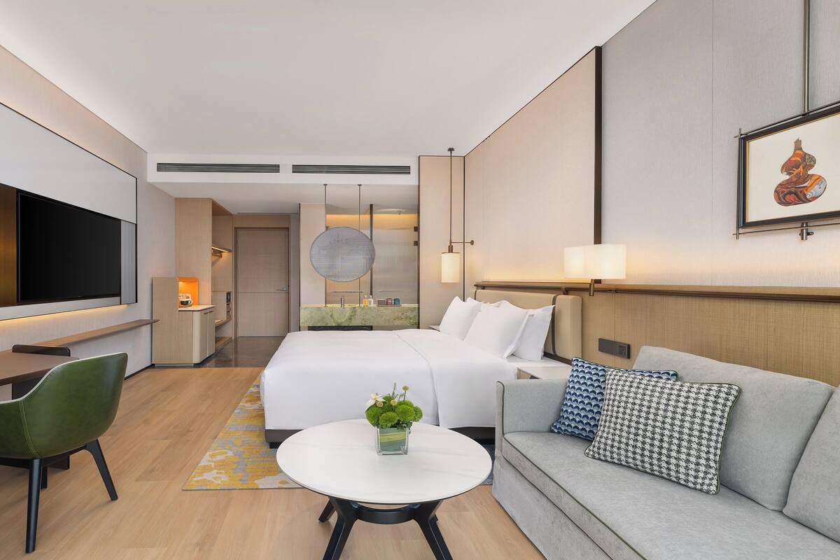 Quarto de hotel moderno com cama, sofá, mesa de jantar, TV e banheiro visível por uma porta aberta.