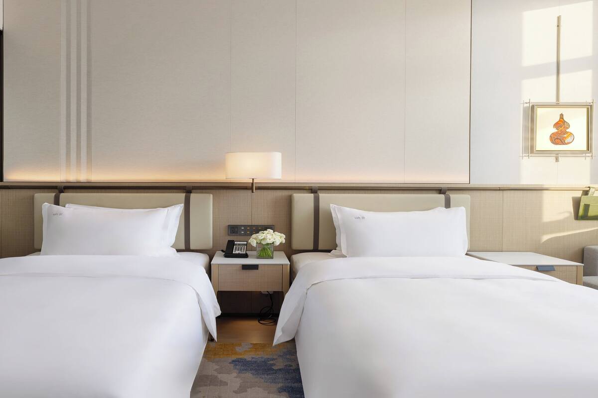 Quarto de hotel com duas camas, uma mesinha de cabeceira com abajur, telefone e um vaso de flores.
