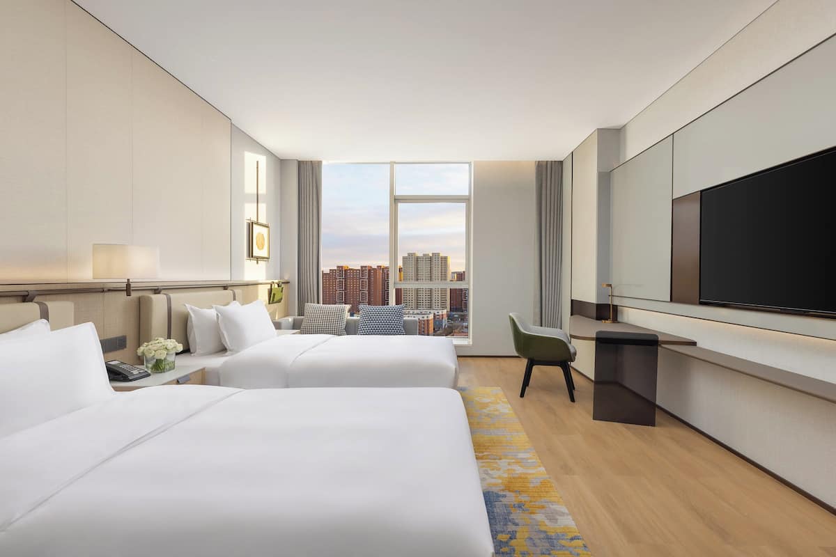 Quarto de hotel moderno com duas camas, uma escrivaninha, TV de tela plana e vista para a cidade.