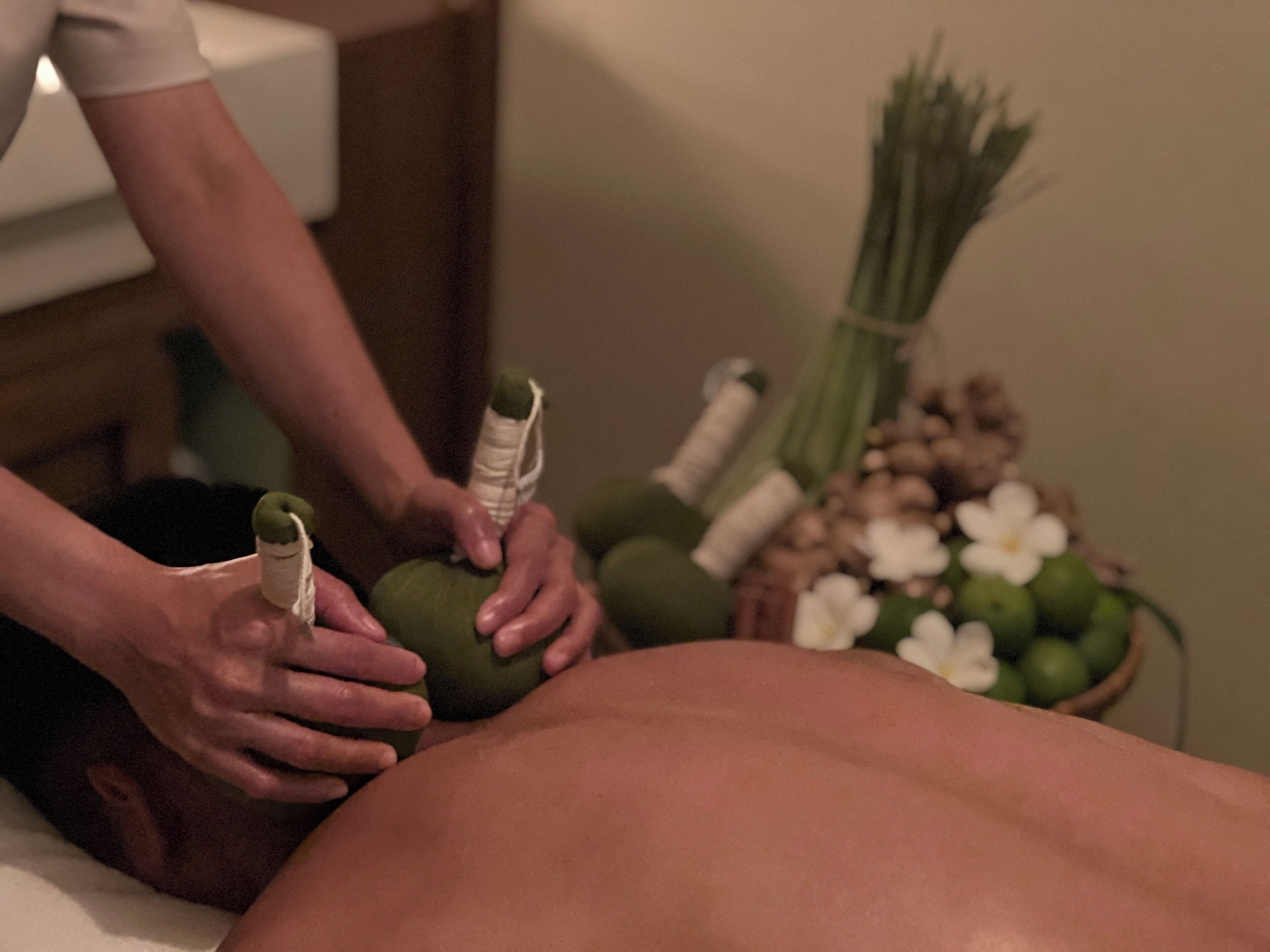 Sessão de massagem com uma pessoa recebendo tratamento nas costas, cercada por diversas ervas e flores.