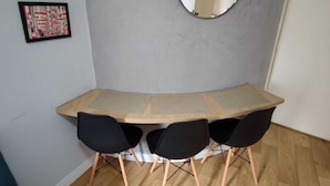 Uma área de refeições no canto, com uma mesa de madeira e cadeiras pretas, um espelho redondo na parede e um quadro emoldurado sobre a mesa.