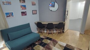 Uma sala de estar com um sofá verde-azulado, um tapete estampado, uma escrivaninha com duas cadeiras e uma parede com quadros.