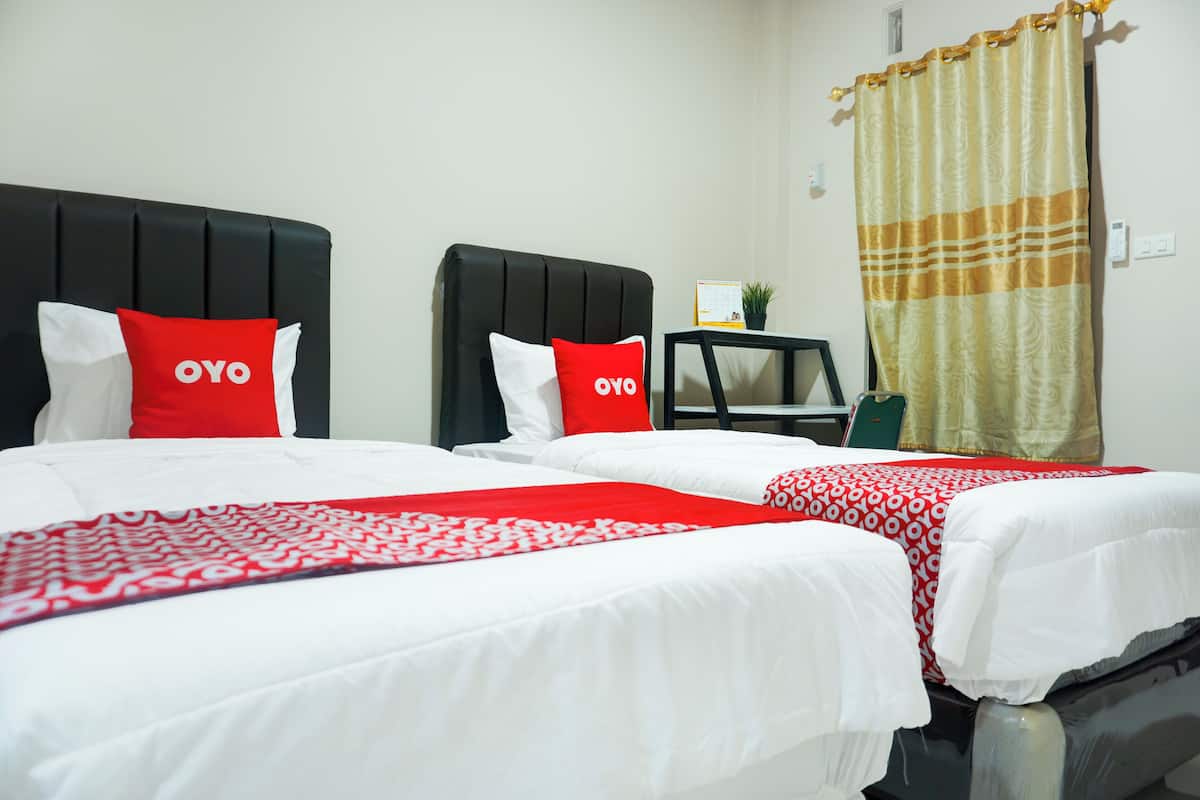 Quarto de hotel com duas camas, cada uma com cabeceira preta e roupa de cama branca. Há almofadas vermelhas com a palavra “OYO” estampada nelas. Uma pequena mesa com uma planta e um calendário é visível no canto.
