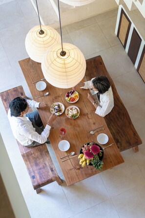 Uma mesa de jantar com três pessoas sentadas, rodeada por pratos de comida, uma saladeira e uma garrafa de bebida vermelha.