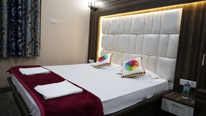 Quarto de hotel com uma cama grande, manta vermelha, dois travesseiros com desenho de flores coloridas, cabeceira de madeira e mesa de cabeceira com garrafa de água.
