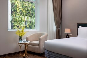 Quarto de hotel com uma cama, uma cadeira, uma pequena mesa com um vaso de flores e uma janela com vista para árvores.