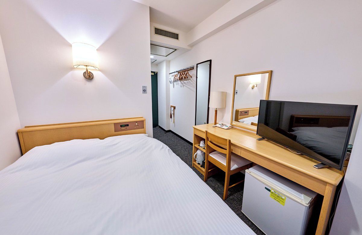 Quarto de hotel com cama, escrivaninha, computador, televisão e pequeno frigorífico.