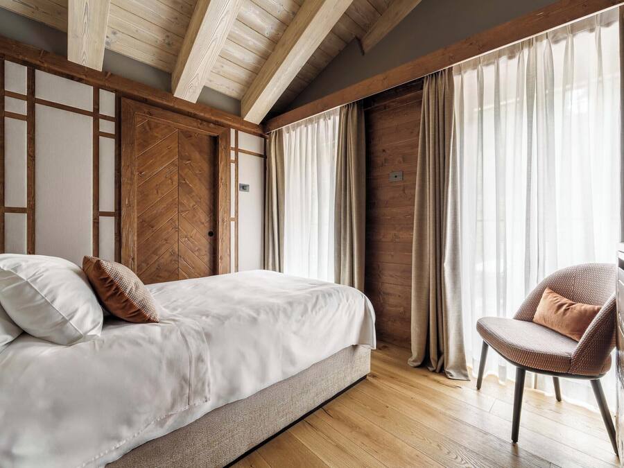 Quarto com cama, cadeira, vigas de madeira e cortinas.