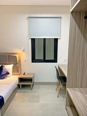 Quarto pequeno e bem iluminado com uma cama, mesa de cabeceira e escrivaninha com cadeira. Há uma janela com persiana e uma tomada na parede.