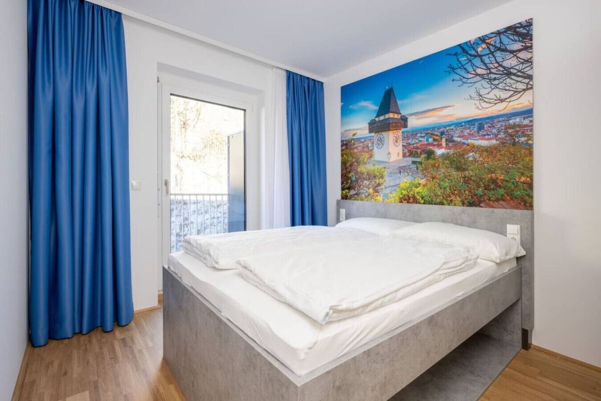 Quarto com cama, janela e pintura de paisagem urbana na parede.