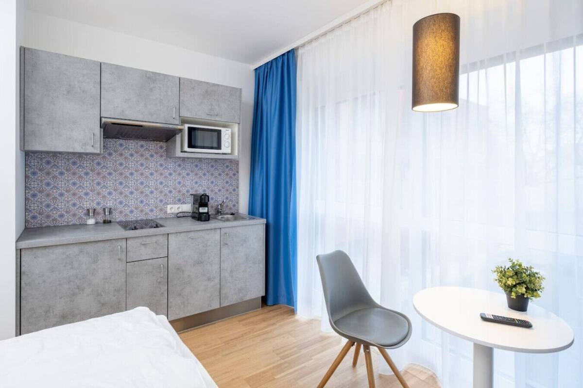 Quarto de hotel moderno com cozinha compacta, uma cama, mesa de jantar com cadeira e janela com cortinas translúcidas.