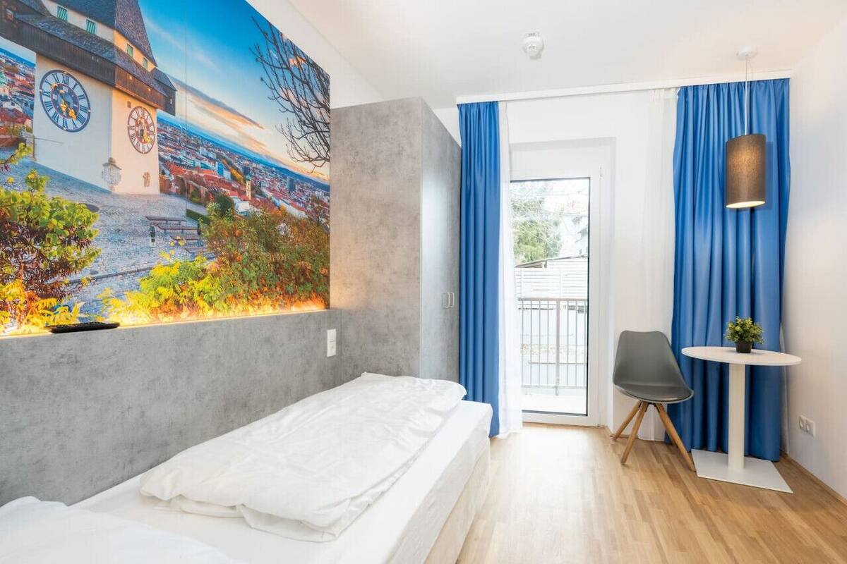 Quarto de hotel moderno com uma cama, um quadro de uma paisagem urbana, uma varanda com cortinas azuis e uma pequena mesa com uma planta.