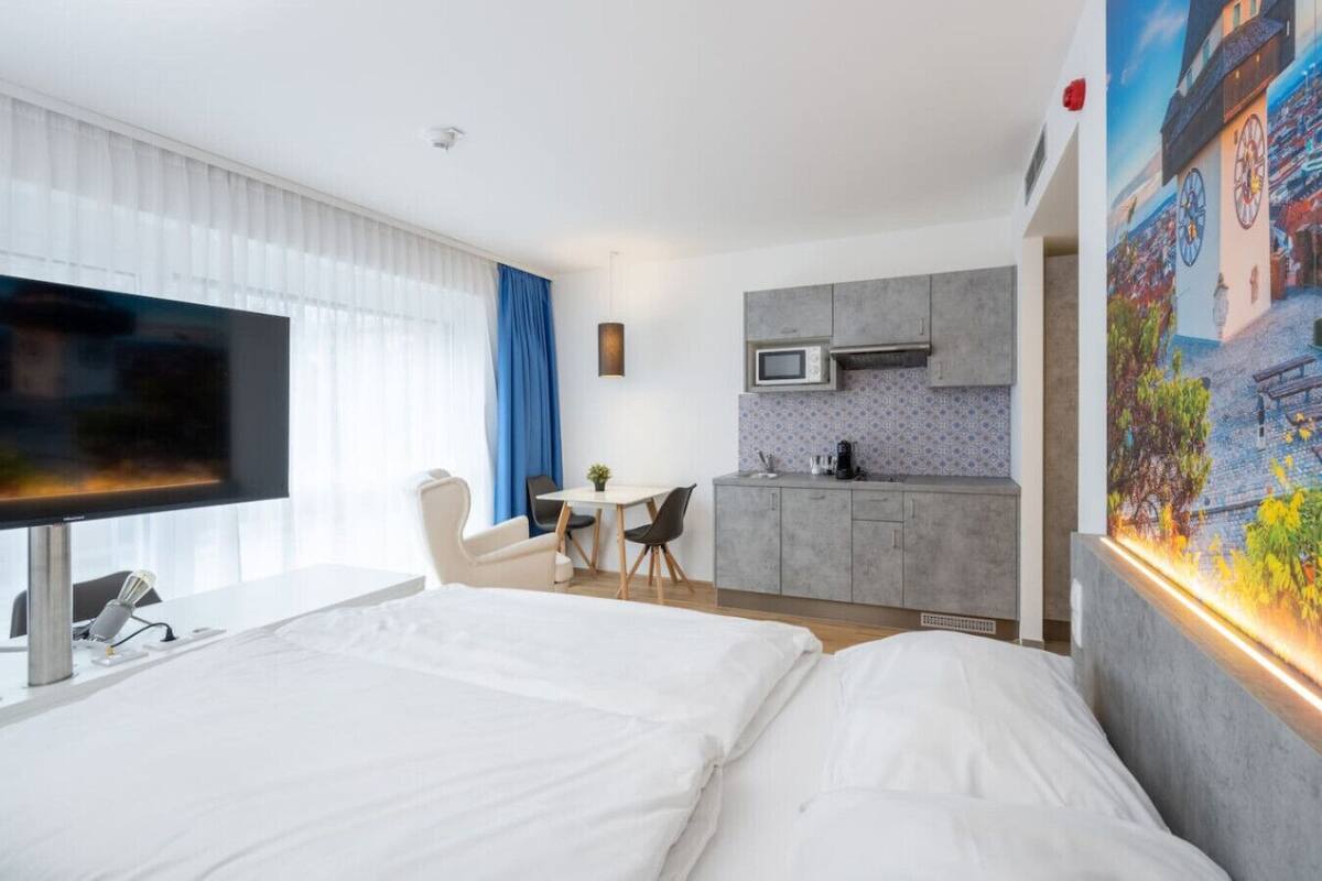 Quarto de hotel com uma cama grande, uma escrivaninha com cadeira, cozinha compacta e televisão.