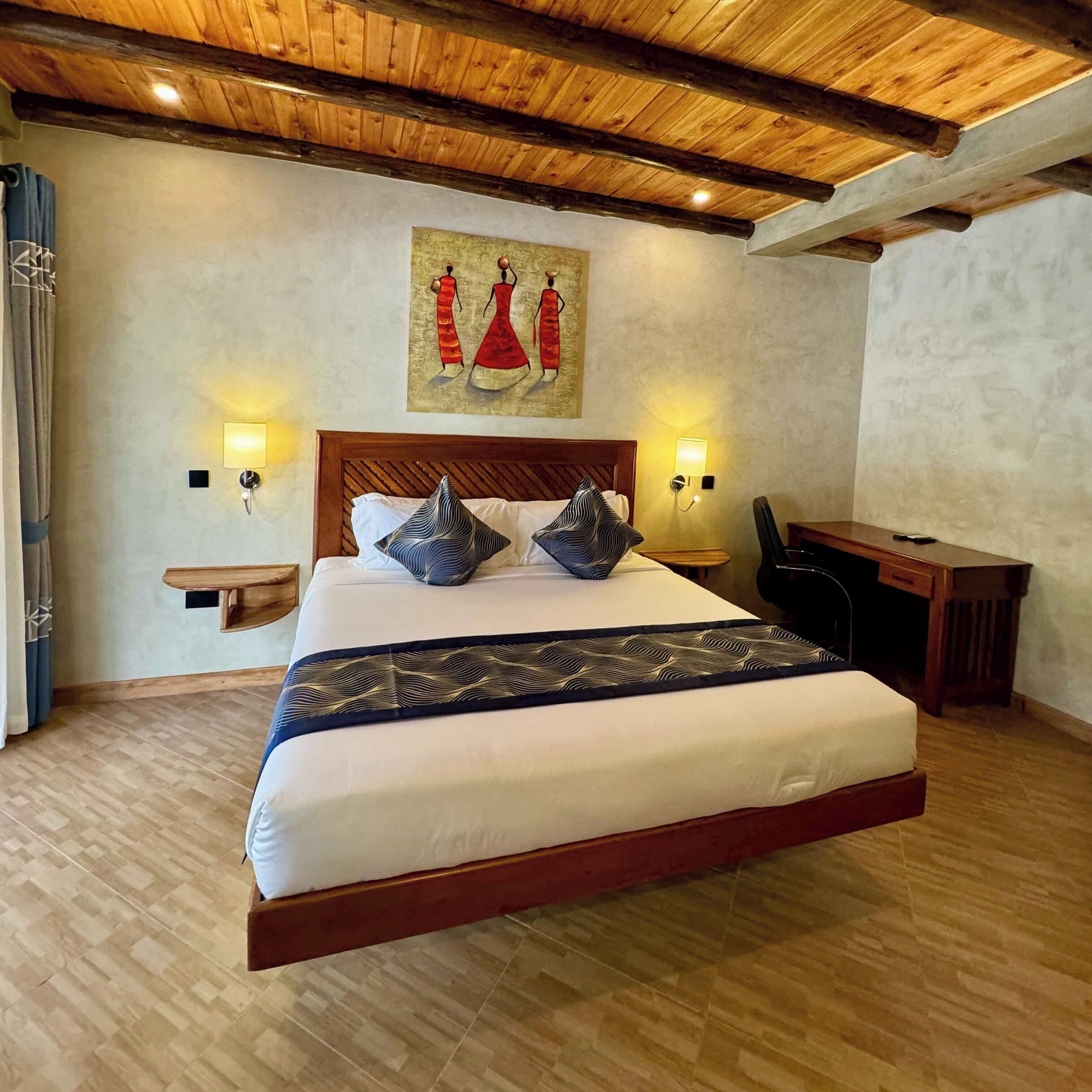 Quarto de hotel com uma cama grande, cabeceira de madeira, duas mesas de cabeceira com luminárias, uma escrivaninha e um quadro na parede.