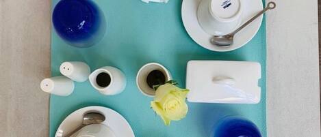 Uma mesa bem organizada com um jogo americano verde-água, apresentando uma chaleira branca, xícaras, pires, um guardanapo enrolado e uma rosa amarela.