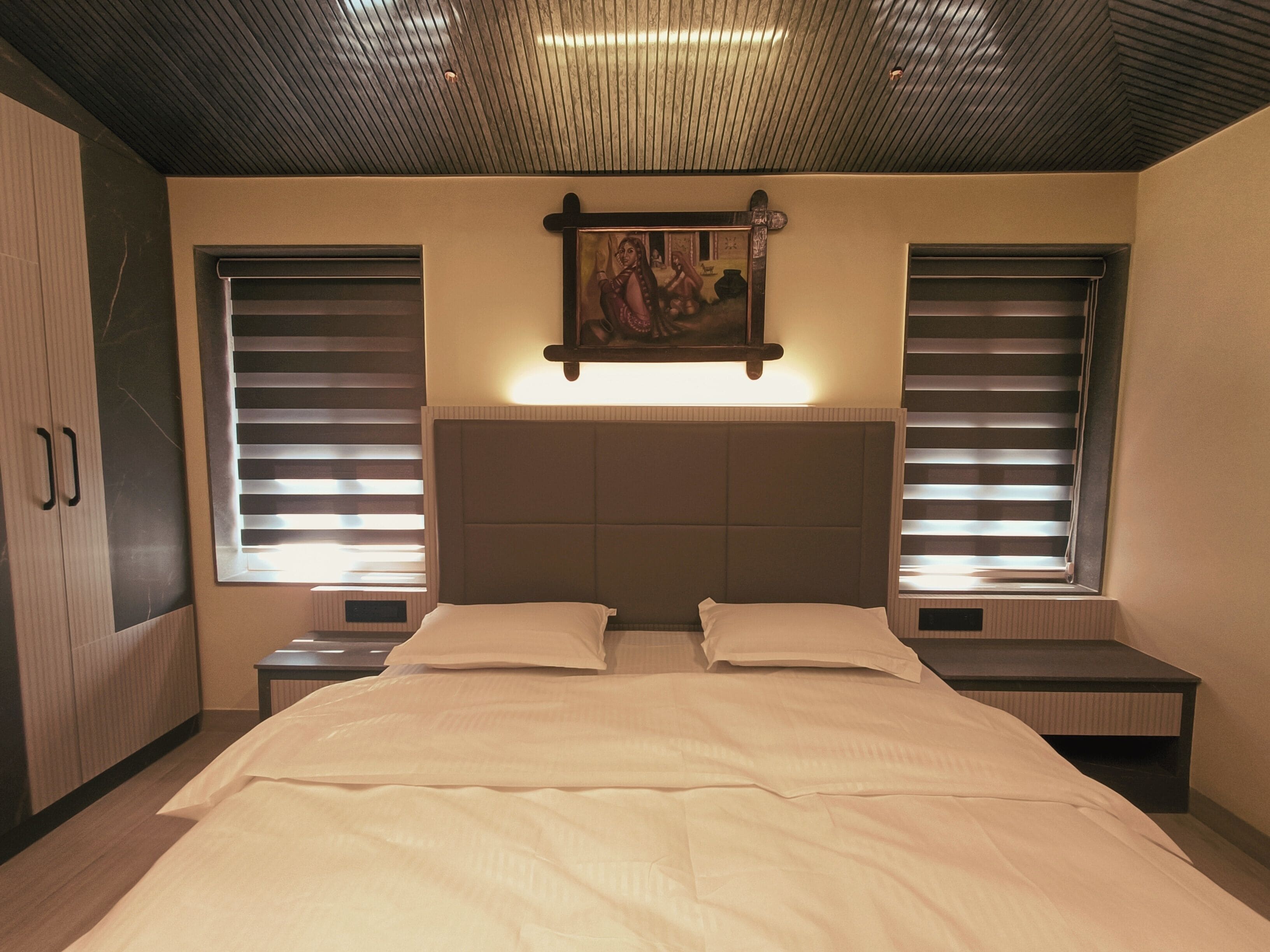 Quarto com uma cama grande, duas mesas de cabeceira, uma cabeceira e duas janelas com persianas.