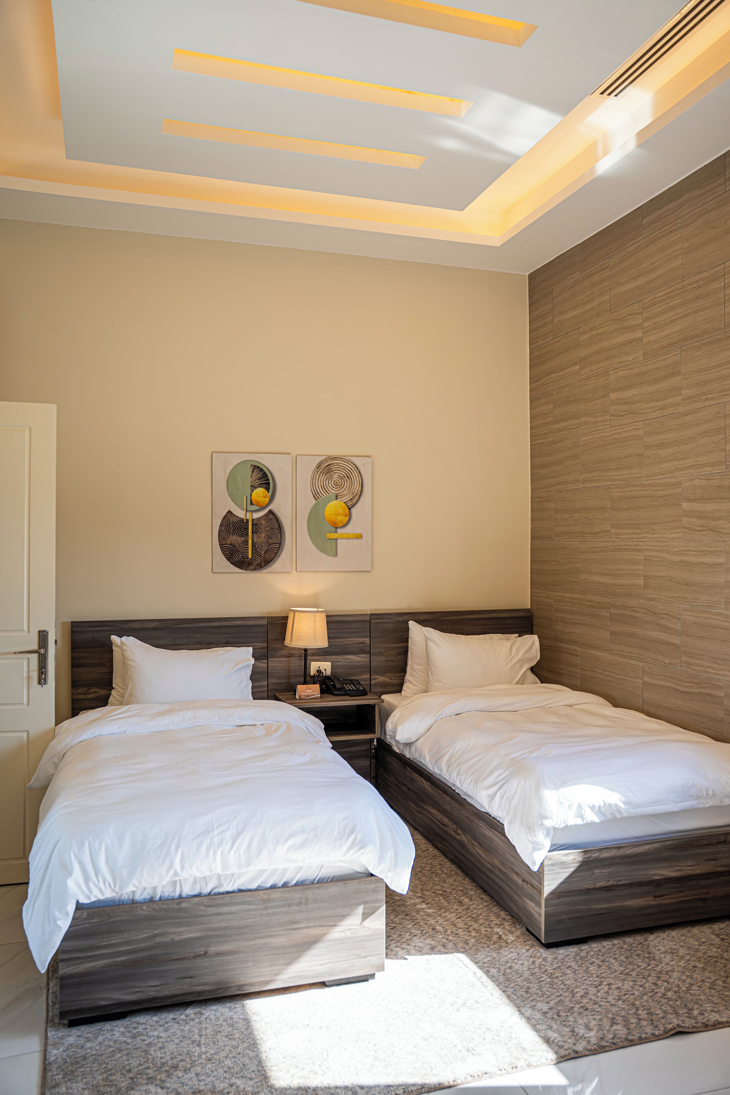 Quarto de hotel moderno com duas camas, uma mesinha de cabeceira com abajur e uma obra de arte na parede.