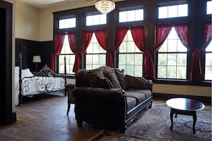Quarto com sofá de couro marrom escuro, poltrona combinando e uma mesa redonda pequena. Janelas amplas com cortinas vermelhas permitem a entrada de luz natural.