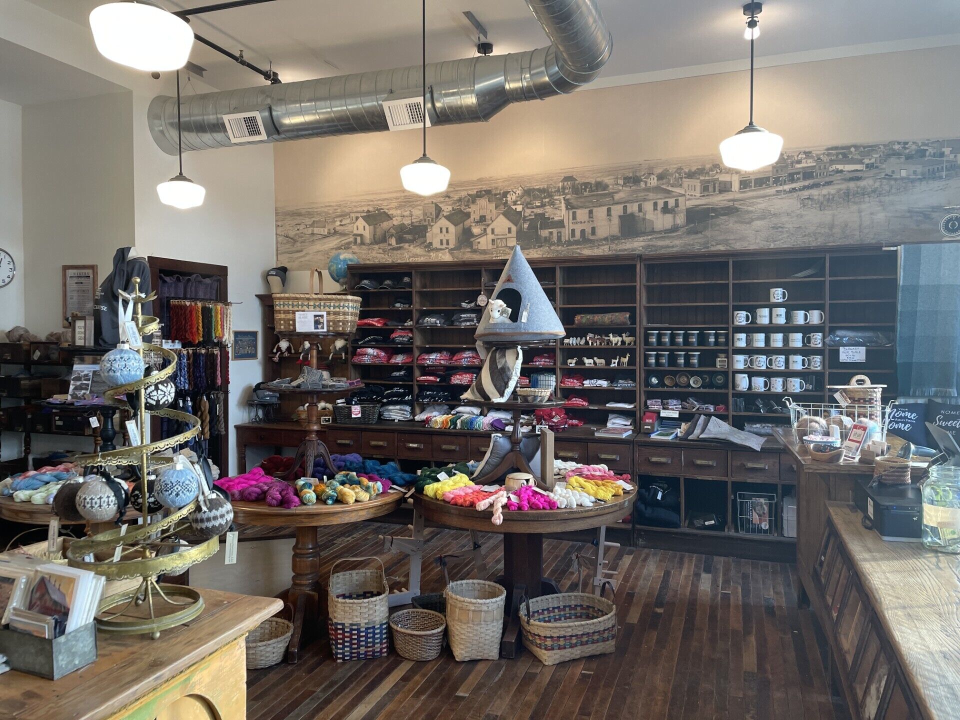 Uma boutique acolhedora com prateleiras de lãs coloridas, diversos sacos e um mural antigo de paisagem urbana.