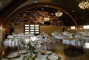 Salão de banquetes com várias mesas redondas preparadas para refeições, com toalhas brancas, arranjos de mesa verdes e detalhes dourados.