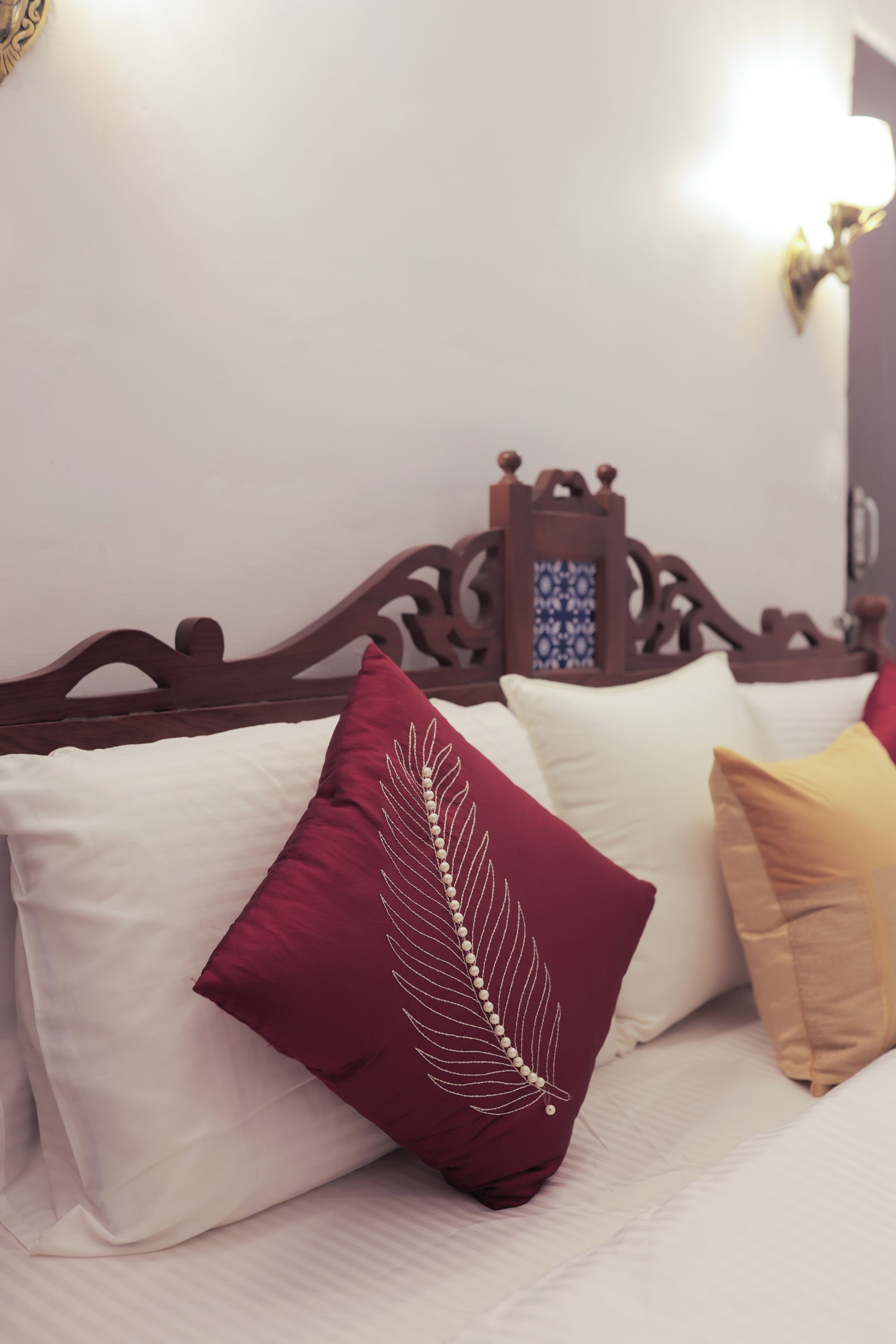 Uma cama com cabeceira de madeira, almofadas brancas e borgonha, e um candeeiro de parede decorativo.