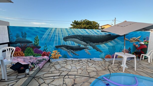 Um pátio externo com um mural de baleias e corais, piso de pedra e cadeiras de plástico brancas.