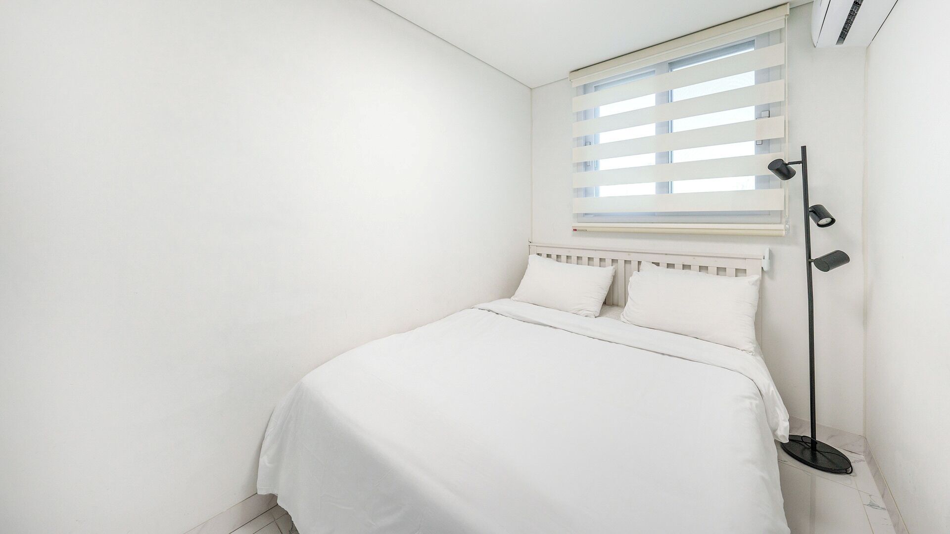 Um quarto claro com uma cama, uma janela com persianas e um abajur.