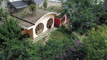 Um pequeno edifício térreo com telhado inclinado, uma porta vermelha e um jardim com diversas plantas e um caminho.