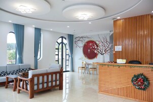 Uma área de recepção de hotel com um balcão de madeira, um espaço de estar com sofás e cadeiras, e amplas janelas com cortinas azuis.