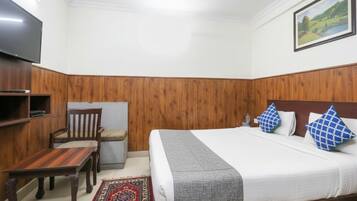 Quarto de hotel com parede revestida de madeira, uma cama com roupa de cama branca e almofadas estampadas em azul, uma televisão à esquerda e uma pequena mesa com uma cadeira.