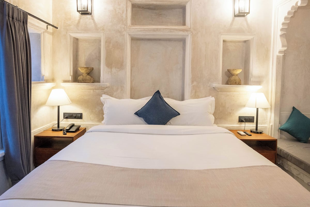 Quarto de hotel com uma cama grande, duas mesas de cabeceira com abajures, um telefone e objetos decorativos nas mesas laterais.