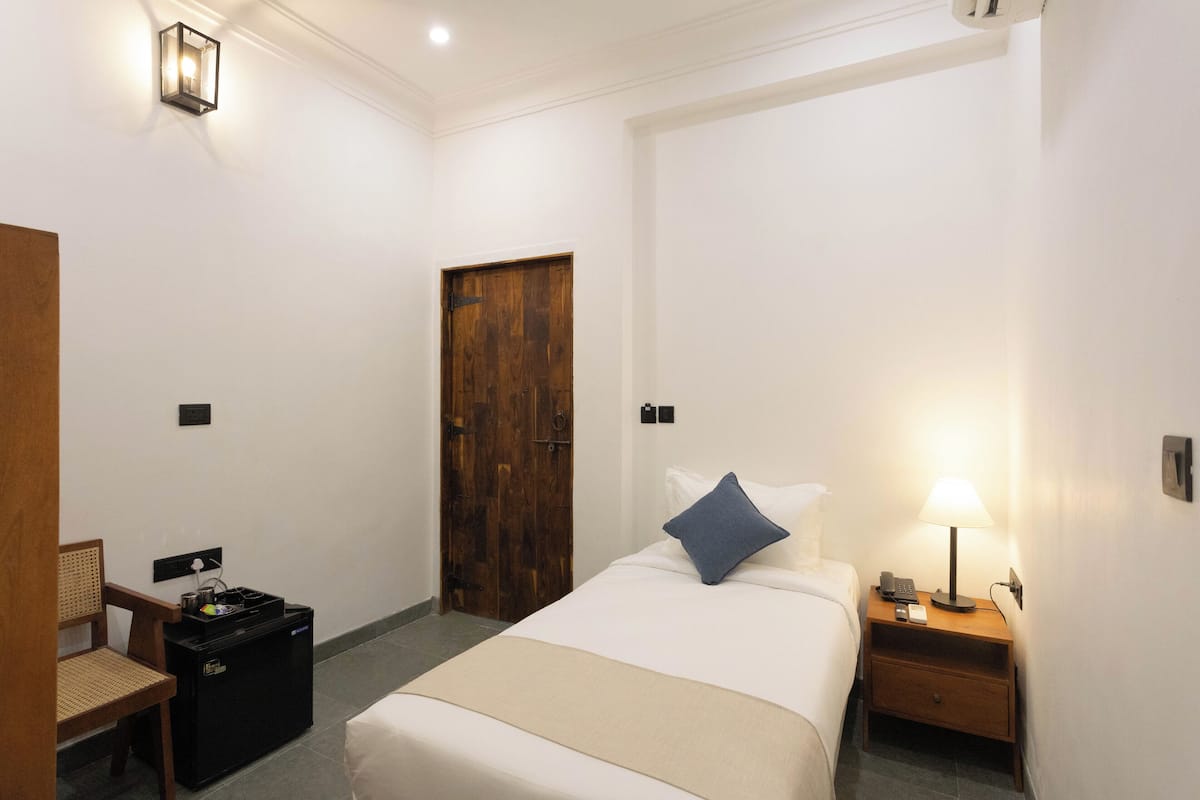 Quarto de hotel com cama, mesa de cabeceira, luminária, cadeira e porta de madeira.