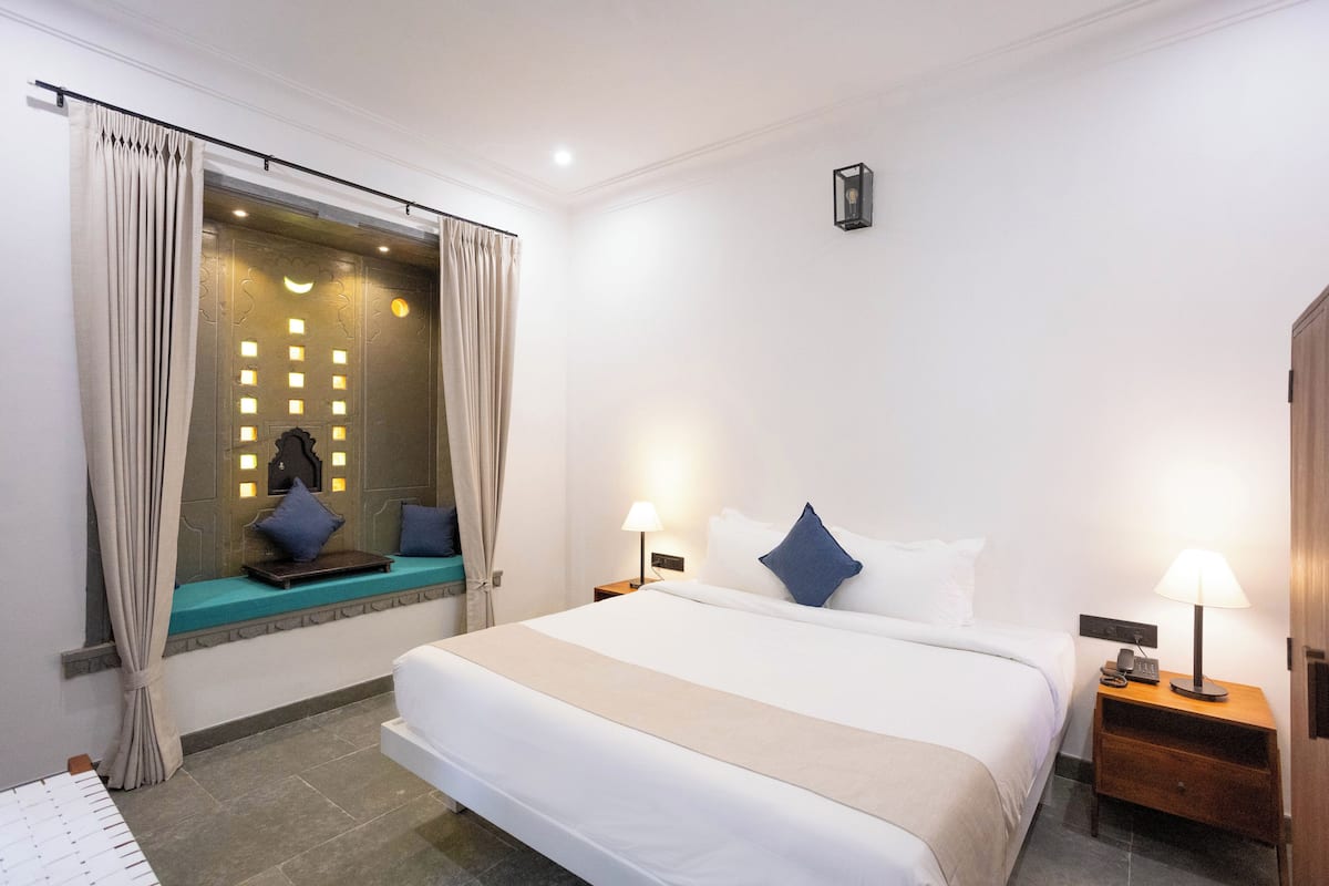 Quarto de hotel com cama, janela com cortinas, mesa de cabeceira com abajur e luminária na parede.