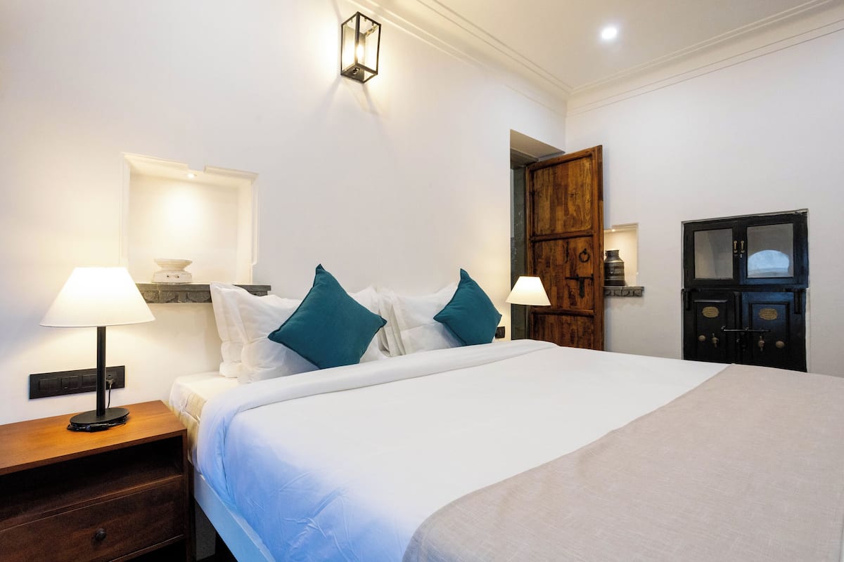 Cama bem arrumada com almofadas brancas e verde-água, abajur e guarda-roupa de madeira num quarto bem iluminado com paredes brancas.