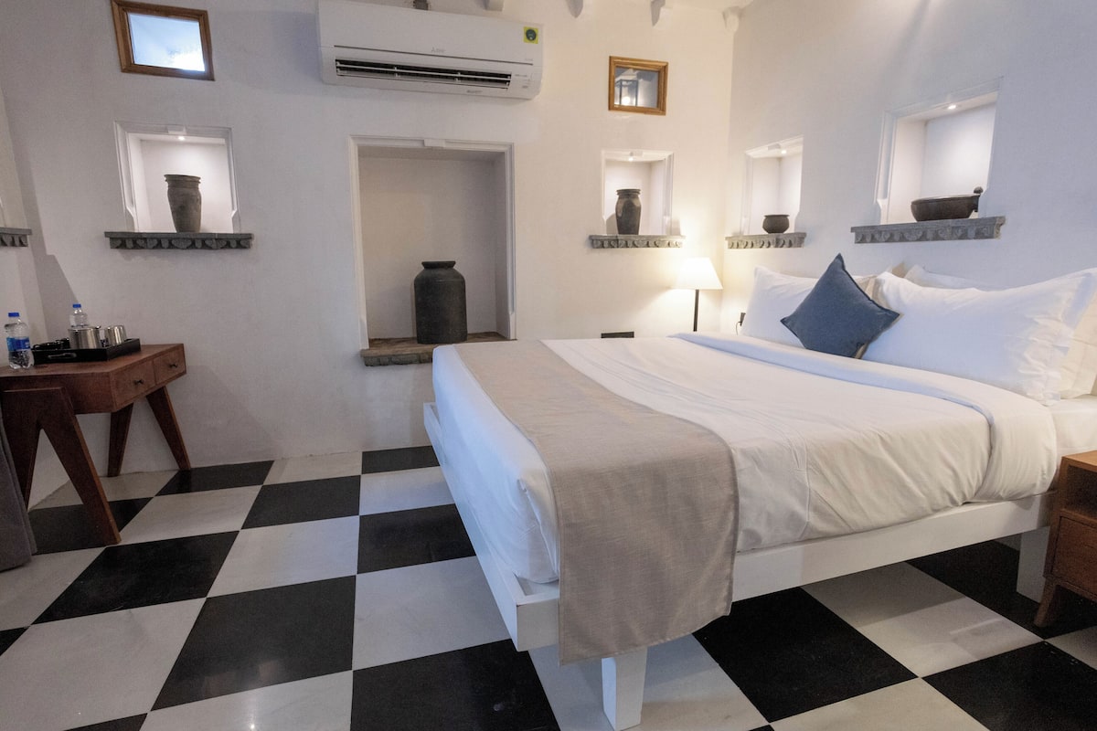 Quarto de hotel com chão quadriculado, cama com roupa de cama branca, mesa de cabeceira com abajur e prateleiras na parede com objetos decorativos.