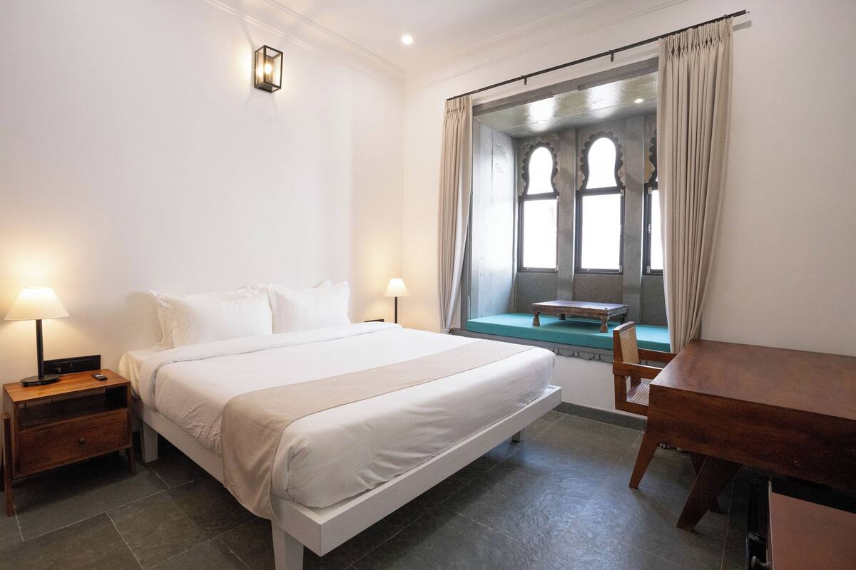 Um quarto com cama, mesas de cabeceira, escrivaninha e janela com cortinas.